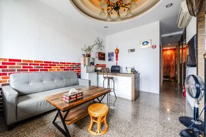 Uma sala de estar moderna com um sofá cinzento, mesa de centro de madeira e um banquinho de vime. Uma área de cozinha com um banquinho de bar e um ventilador pode ser vista ao fundo.