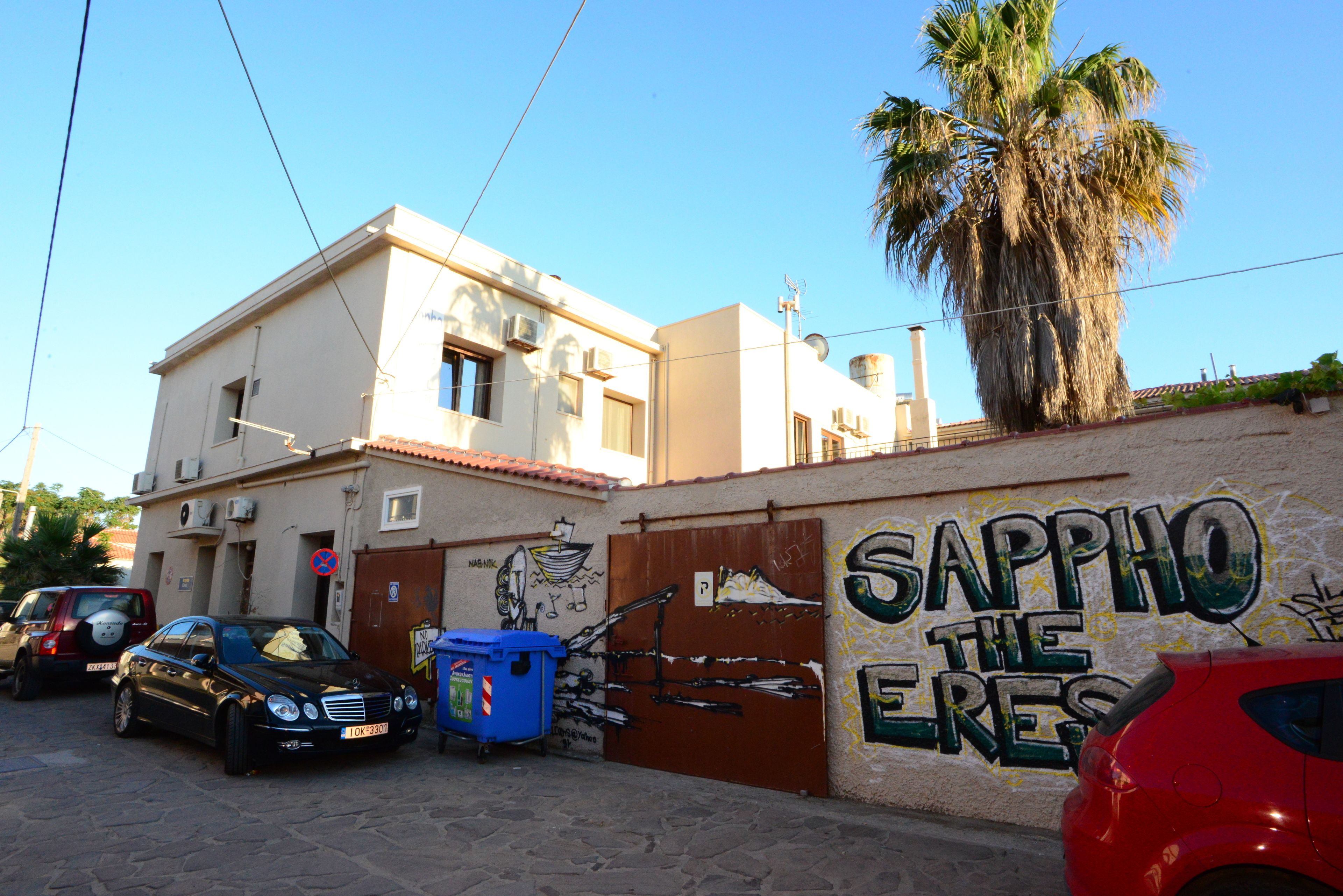 Um edifício com um mural que diz "SAPPHO THE ERES", carros estacionados e uma palmeira.