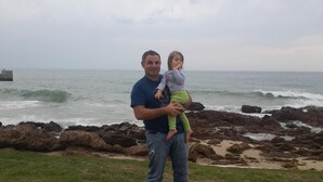 Um homem segura uma criança perto de uma costa rochosa, com ondas quebrando ao fundo.