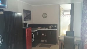Cozinha compacta com geladeira, micro-ondas e pia. Há um relógio na parede e um espaço de refeições com uma cadeira.