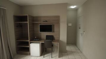 Quarto compacto com escrivaninha embutida, televisão fixada na parede, guarda-roupa e um pequeno frigorífico.