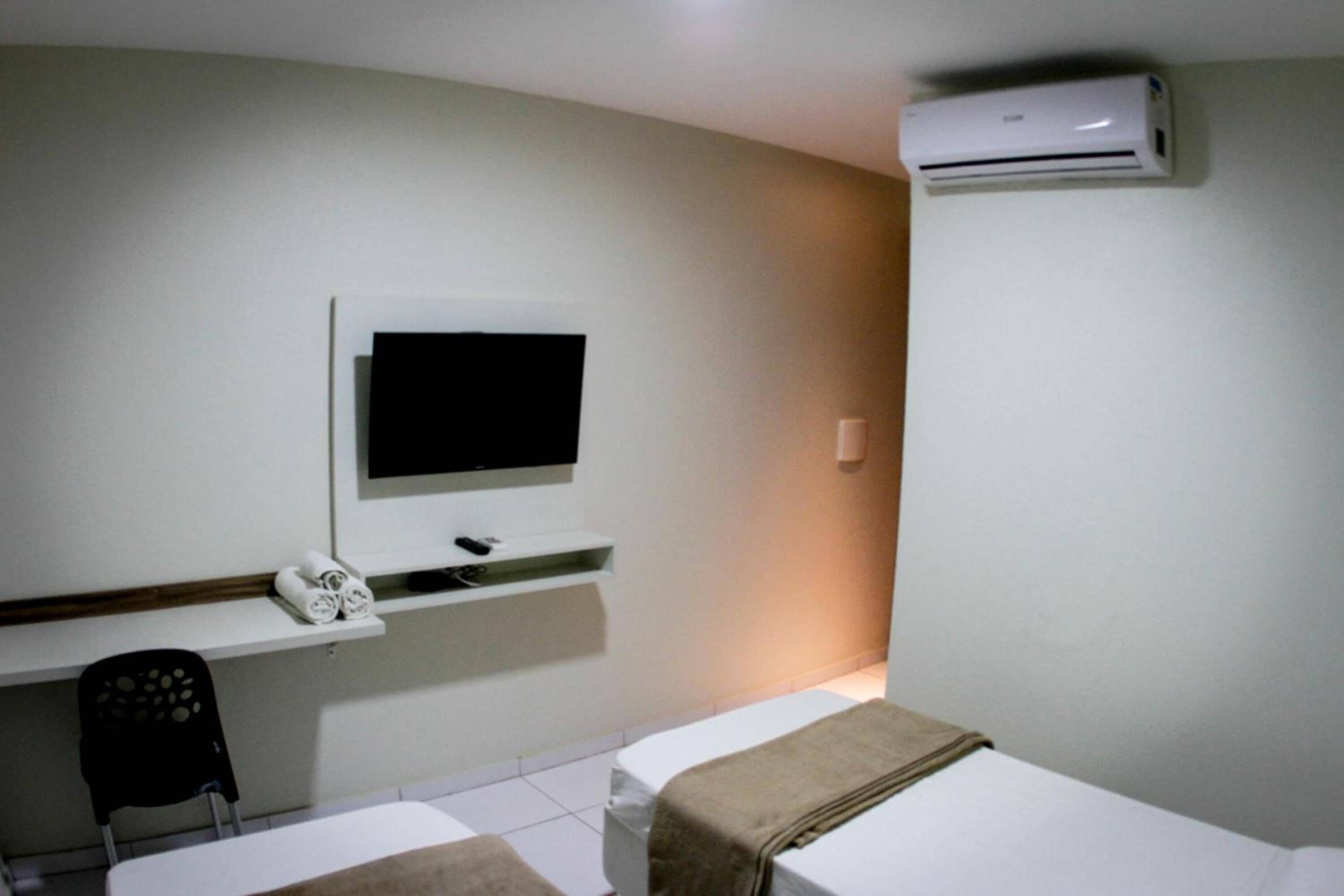 Quarto de hotel com cama, escrivaninha, cadeira, televisão na parede e aparelho de ar condicionado.