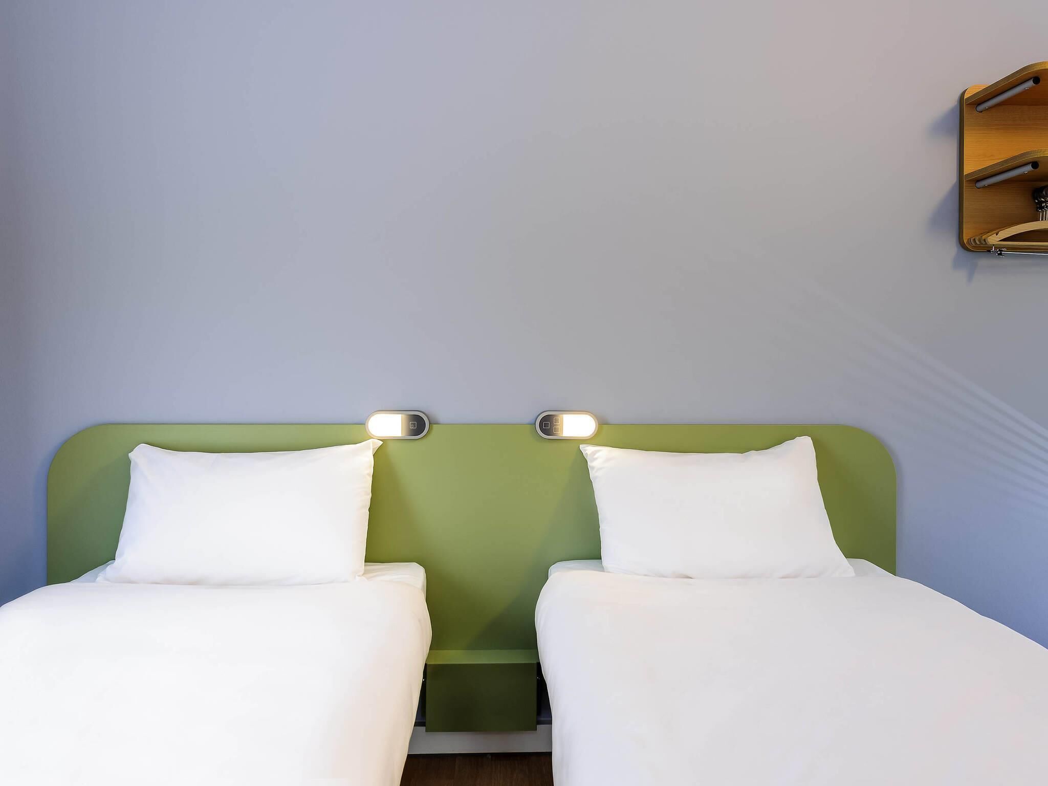 Cama de casal com cabeceira verde e roupa de cama branca, duas mesas de cabeceira com luminárias e uma prateleira na parede.