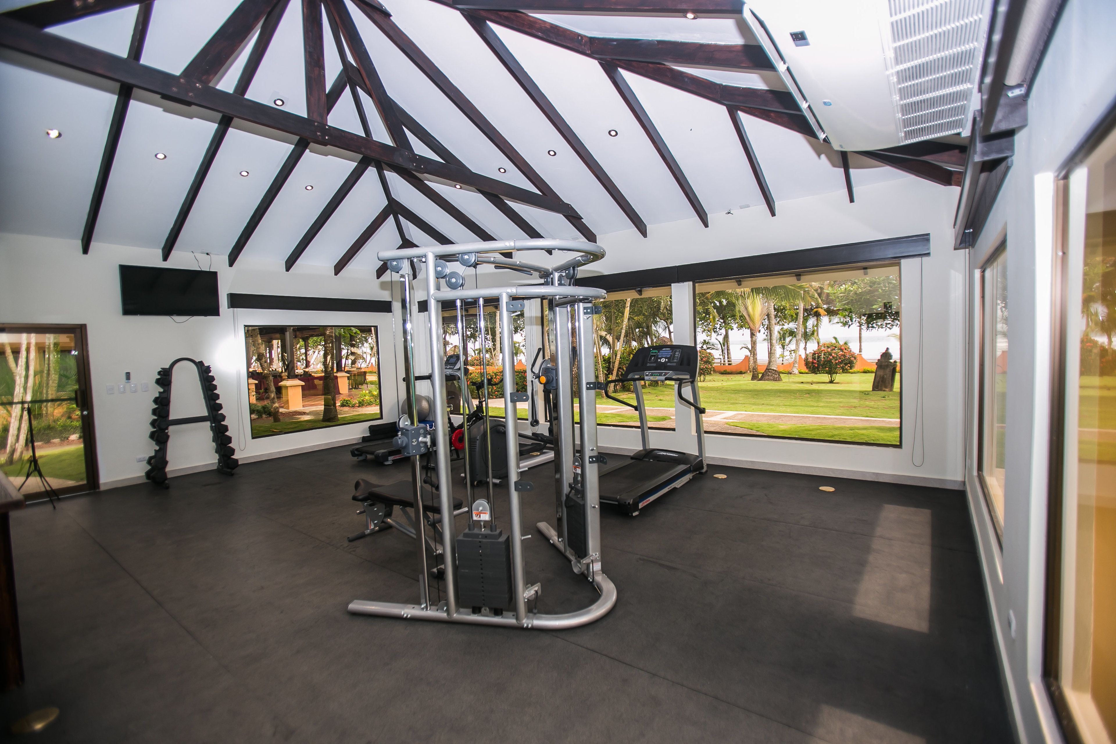 Uma academia com equipamentos de ginástica, grandes janelas e vista para um parque.