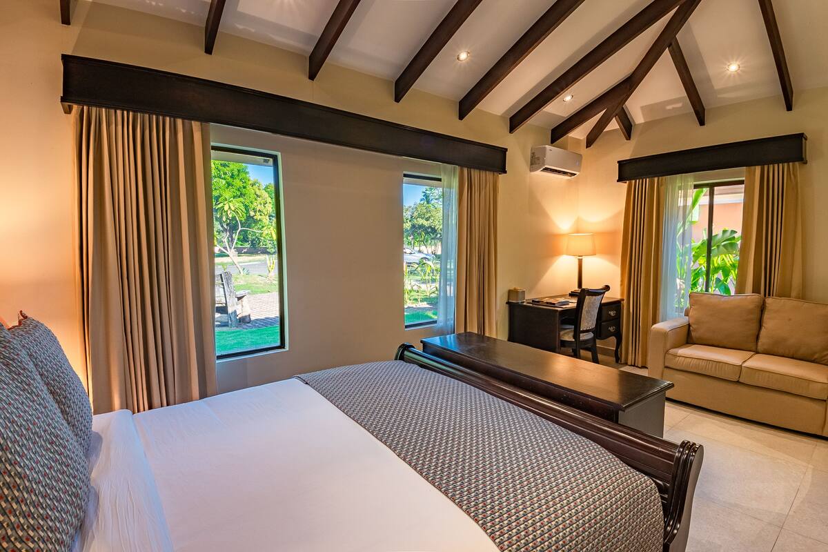 Quarto de hotel com uma cama grande, uma escrivaninha, um sofá e duas janelas com cortinas.