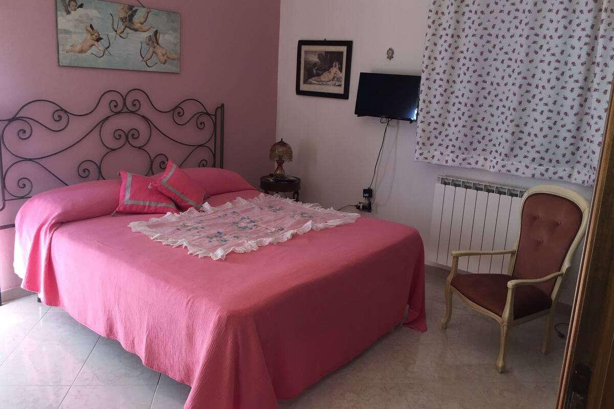 Quarto com cama rosa, uma cadeira, televisão e obras de arte nas paredes.