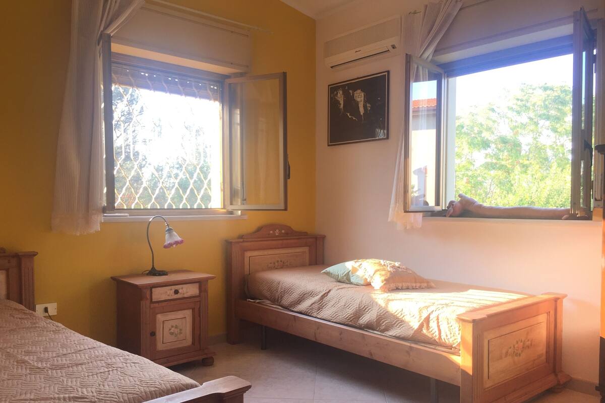 Quarto com duas camas de madeira, uma mesa de cabeceira com abajur e uma janela com cortinas.