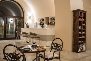 Uma área de refeições com uma mesa posta para dois, dois copos de vinho, um prato de comida e uma pequena tigela decorativa. Há duas cadeiras, uma estante e um armário ao fundo.