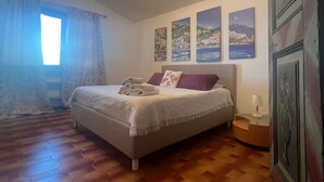 Quarto com cama, mesa de cabeceira, janela com cortinas e três quadros na parede.