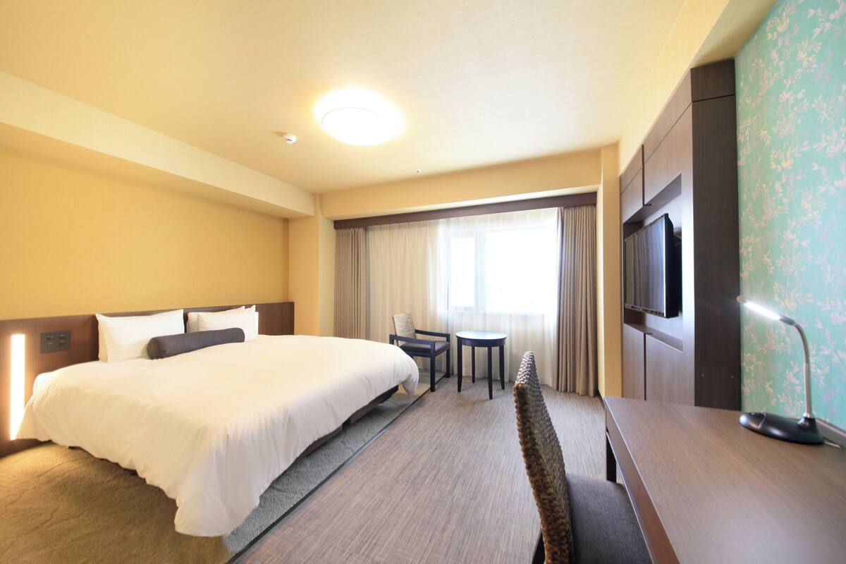 Quarto de hotel com uma cama grande, uma escrivaninha, uma cadeira, uma televisão e uma janela com cortinas.