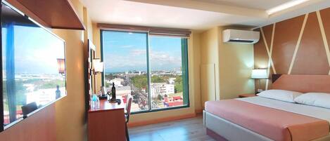 Quarto de hotel com uma janela grande, uma cama, uma escrivaninha e um aparelho de ar condicionado.