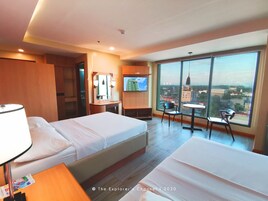 Quarto de hotel com duas camas, televisão, escrivaninha e uma grande janela com vista para a cidade.