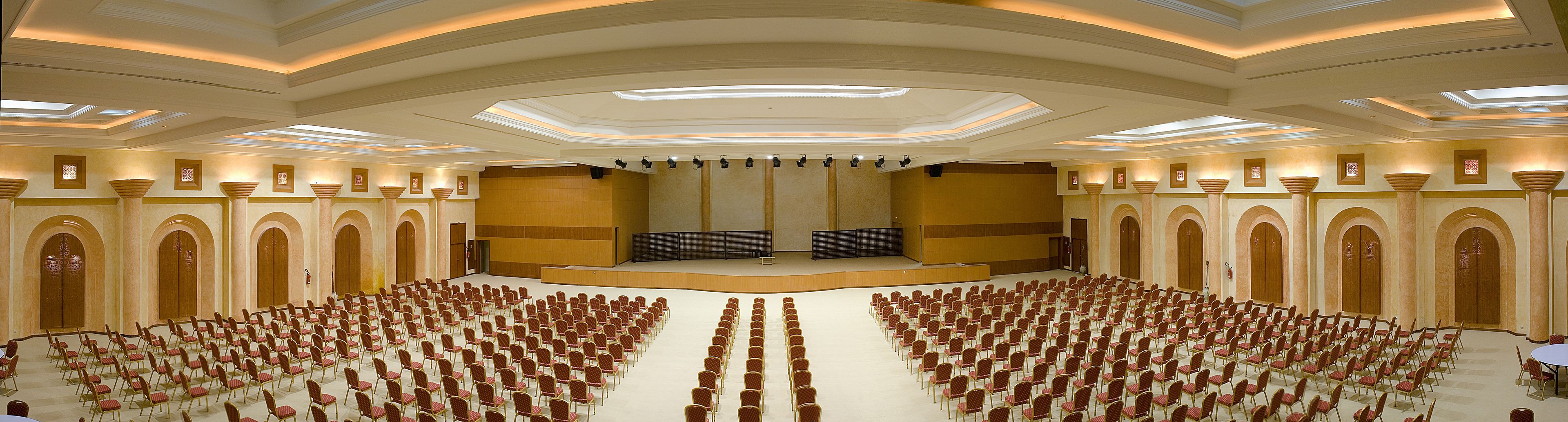Um salão amplo com fileiras de cadeiras voltadas para um palco, portas em arco e um teto alto com claraboias.