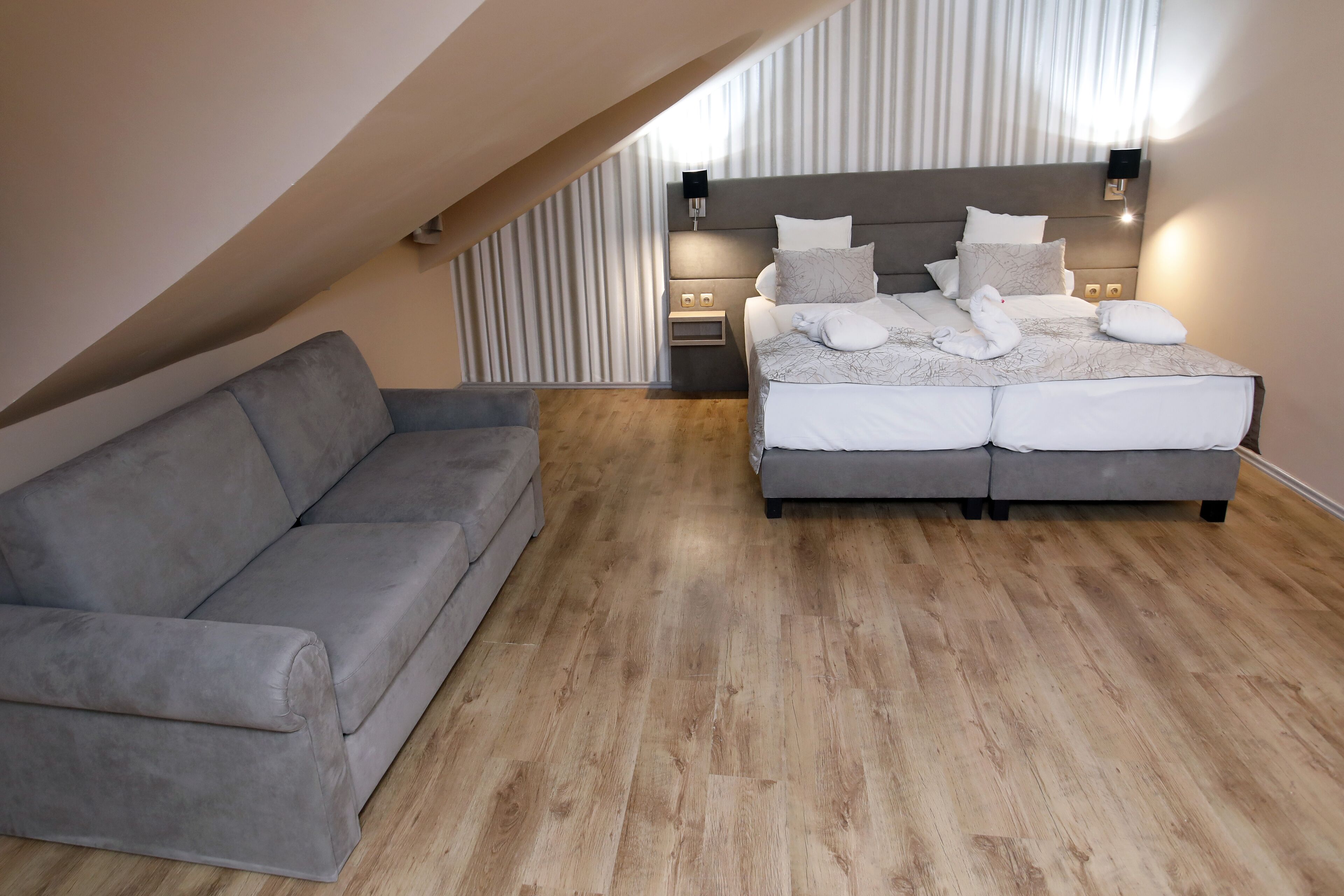 Um quarto moderno com teto estilo loft, um sofá cinzento, uma cama de casal com roupa de cama branca e uma mesa de cabeceira com abajur.
