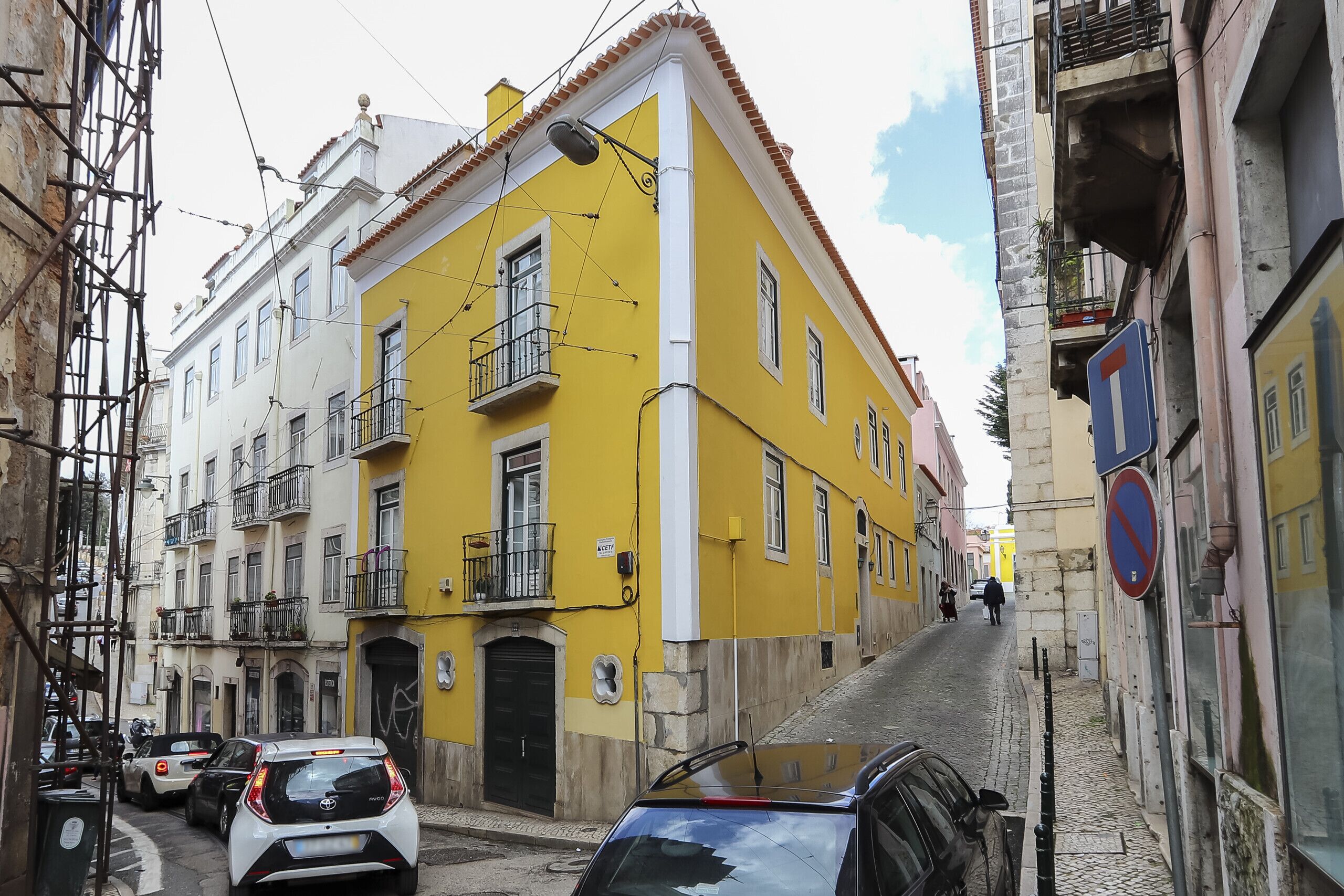 Uma rua estreita com edifícios amarelos e brancos, carros estacionados e um pedestre ao longe.