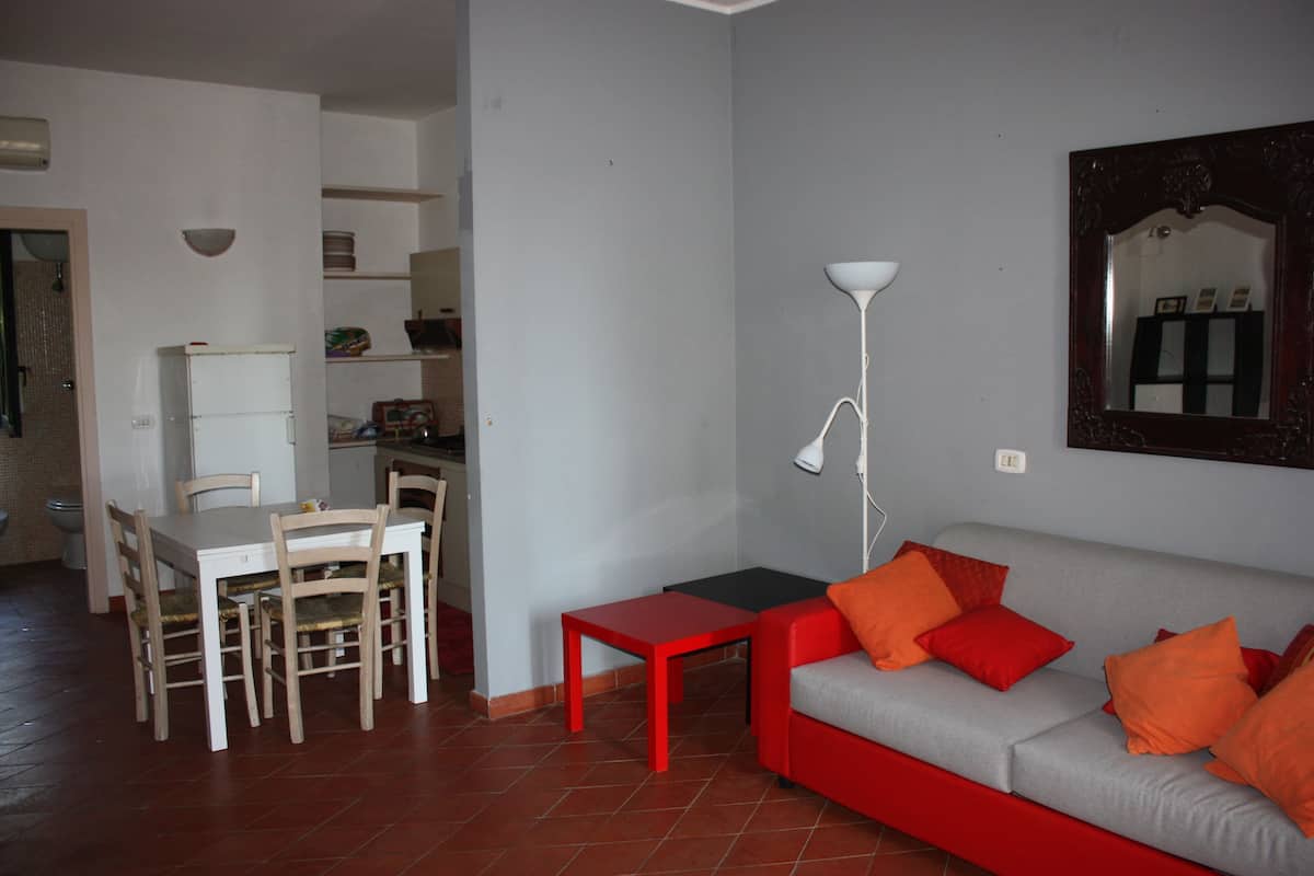Uma sala de jantar com uma mesa e cadeiras brancas, uma mesinha lateral vermelha e um sofá cinza com almofadas laranja.