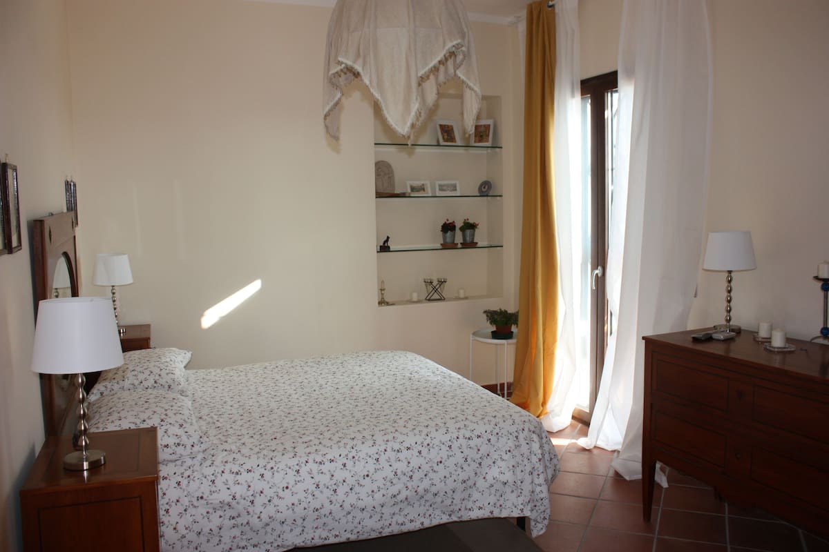 Quarto com cama, mesas de cabeceira, penteadeira, janela com cortinas e prateleira com objetos decorativos.