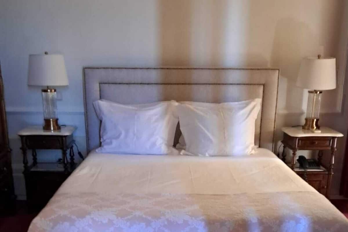Uma cama grande com cabeceira estofada, duas mesas de cabeceira com abajures e um banquinho combinando.