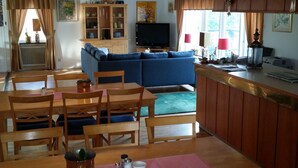 Sala de estar com sofá azul, mesa de jantar de madeira e cadeiras. Há uma televisão e um aparador com objetos decorativos.
