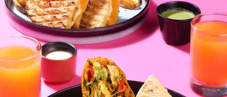 Uma mesa com diversos itens de comida, incluindo sanduíches grelhados, um wrap e uma tigela de batatas fritas, além de copos de suco de laranja.