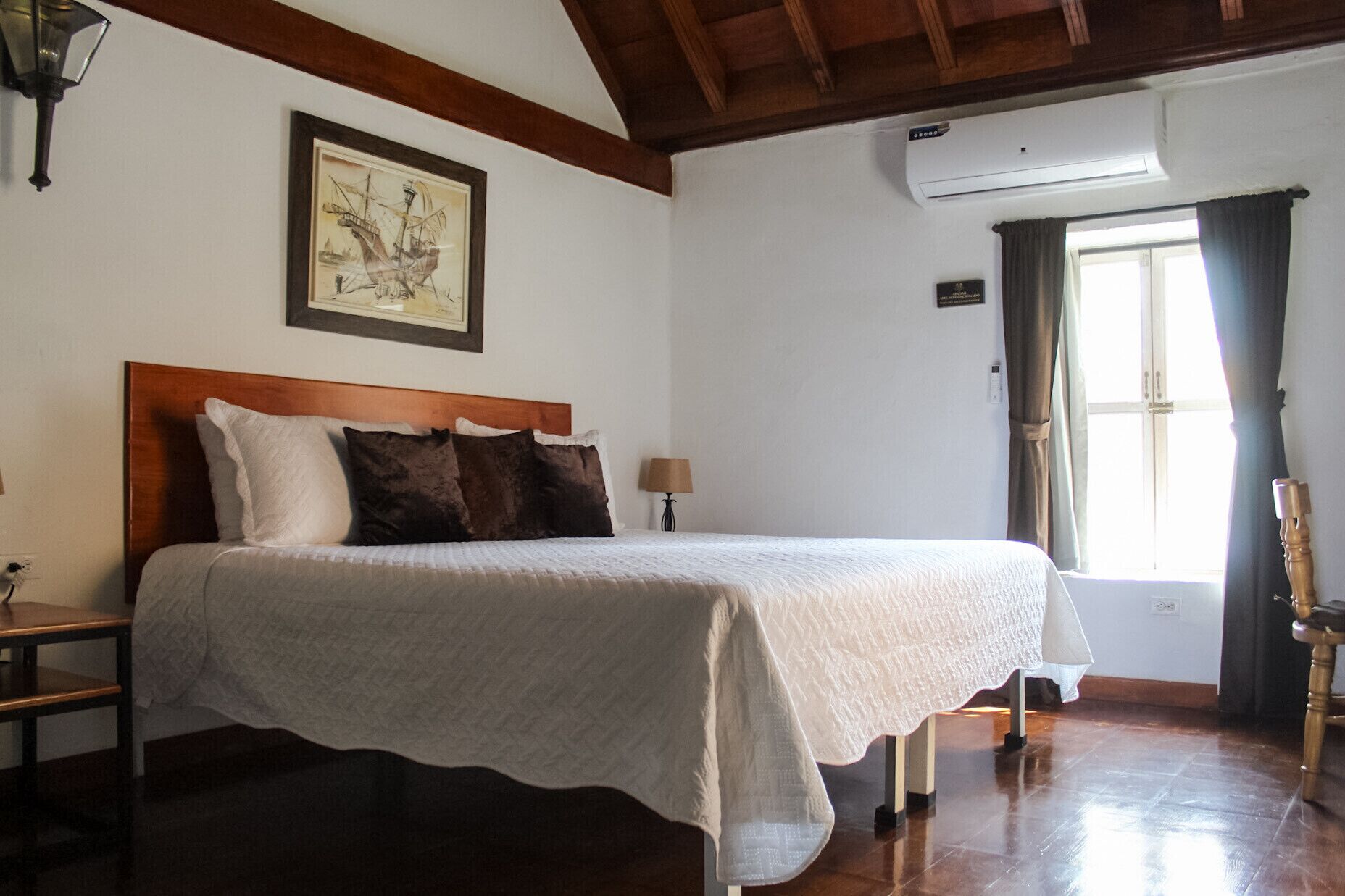 Quarto com uma cama grande, cabeceira de madeira, um quadro na parede, ar condicionado e uma janela com cortinas.