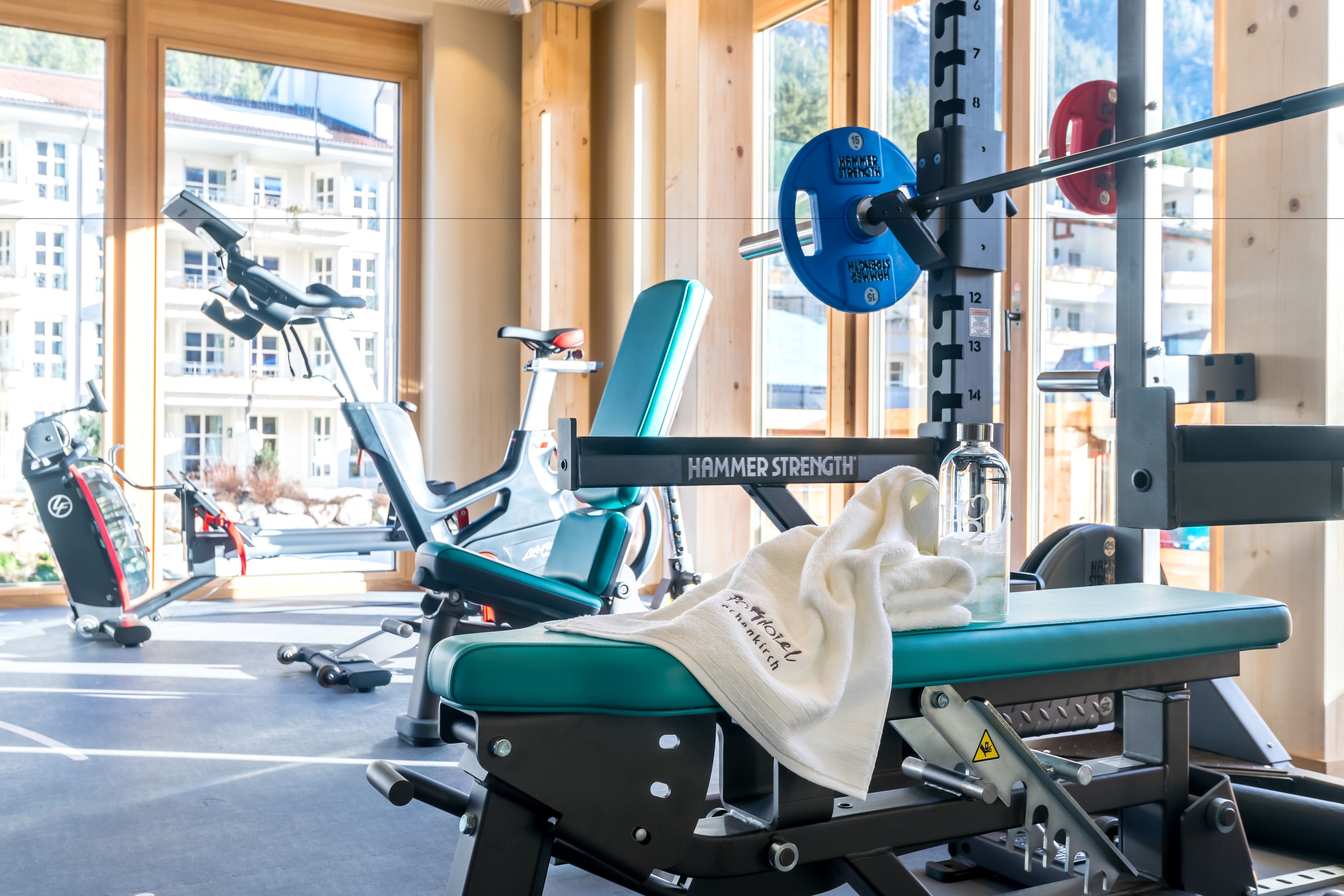 Uma academia com equipamentos de ginástica, incluindo um banco de supino e um banco para pesos, com vista para edifícios lá fora.