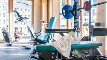 Uma academia com equipamentos de ginástica, incluindo um banco de supino e um banco para pesos, com vista para edifícios lá fora.