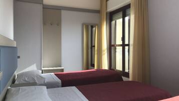 Quarto de hotel com duas camas, um guarda-roupa, uma janela com cortinas e uma pequena mesa.