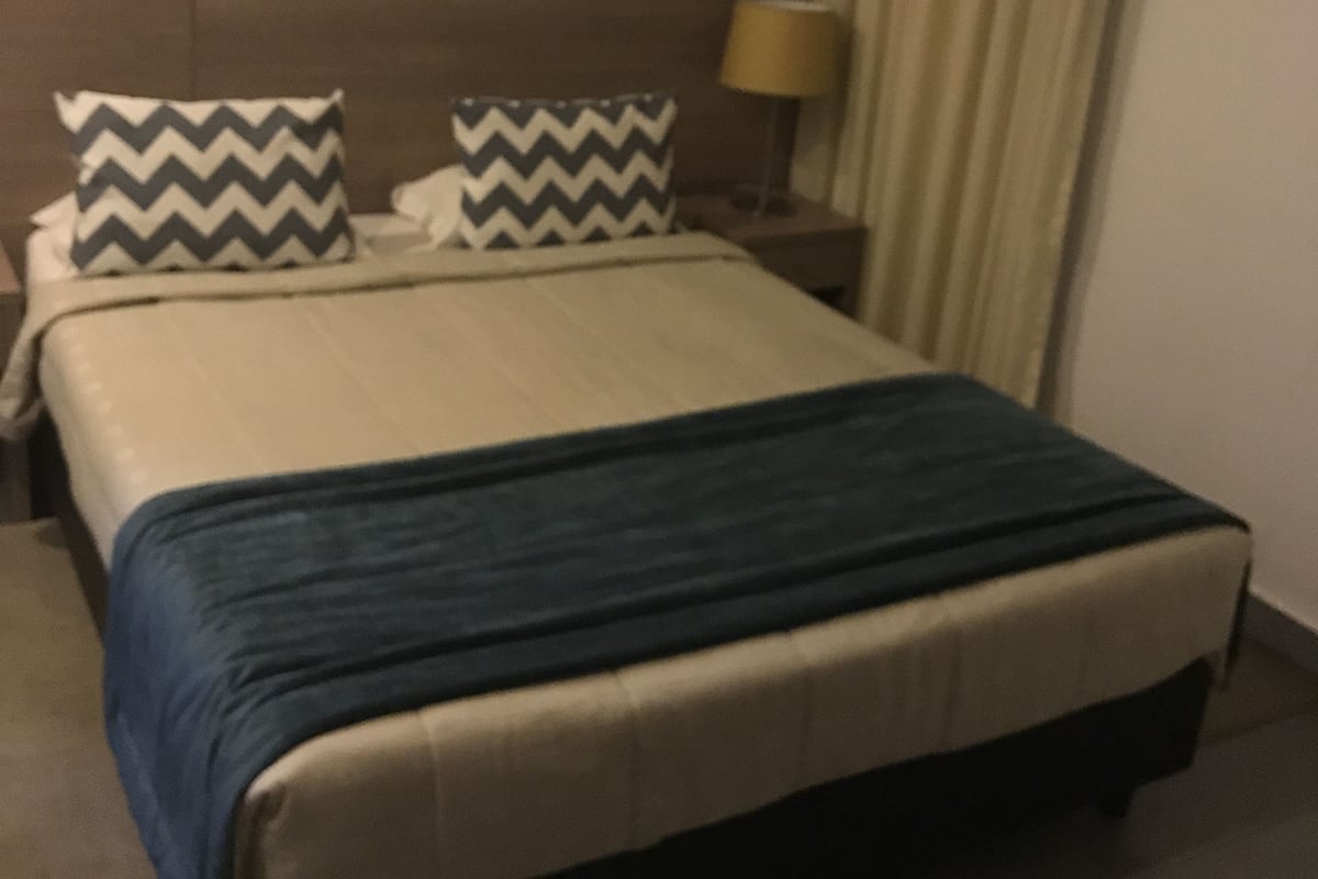 Quarto de hotel com uma cama, duas almofadas, mesa de cabeceira e abajur, e cortina.