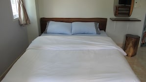 Cama bem arrumada com lençóis brancos e duas almofadas azul claro num quarto com cabeceira de madeira, um banquinho de madeira pequeno e um espelho na parede.