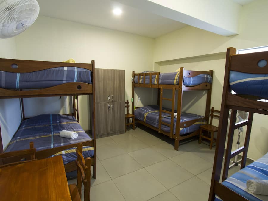 Quarto de dormitório com beliches, uma escrivaninha de madeira, uma cadeira, um guarda-roupa e um ventilador de teto.