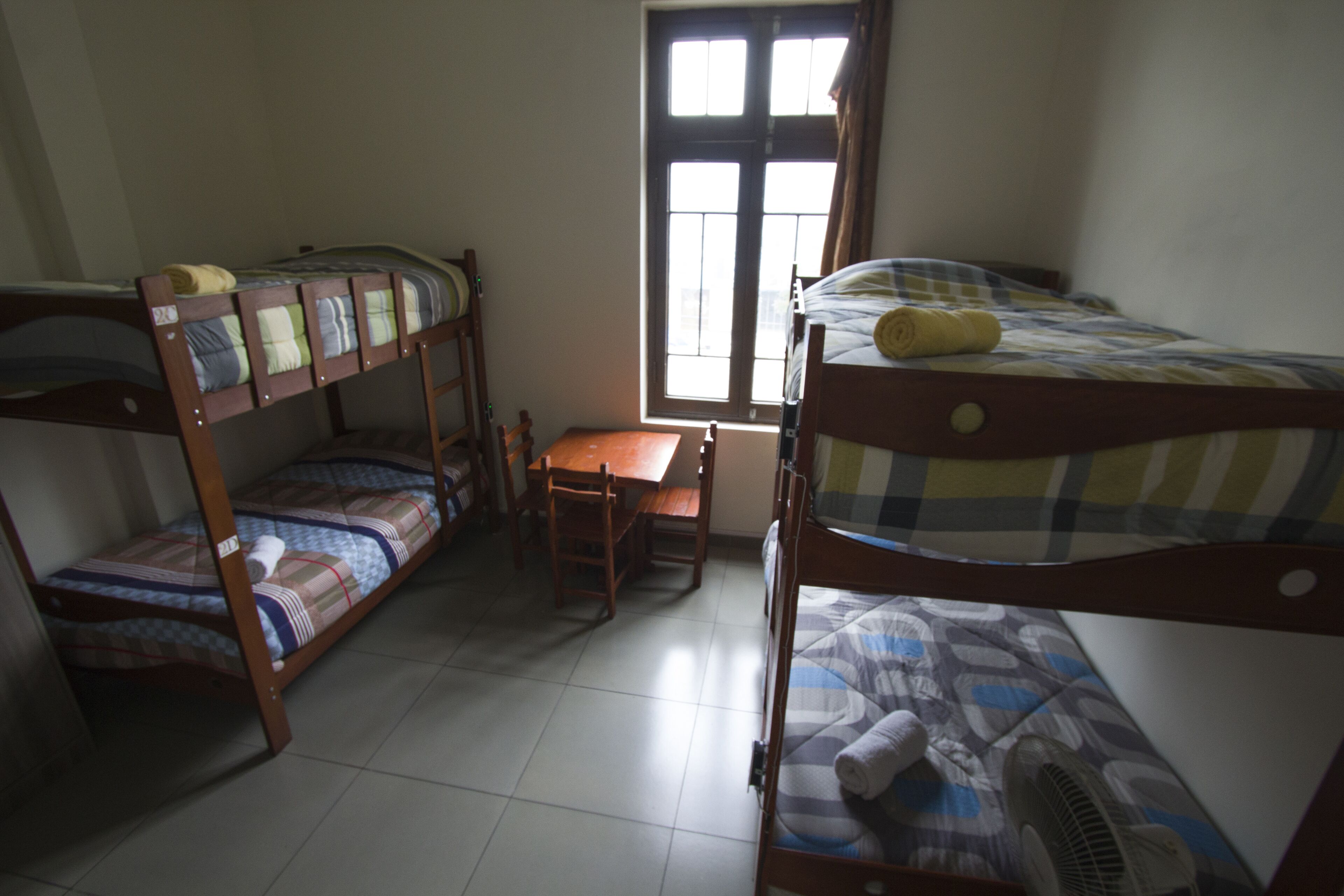 Quarto em dormitório com beliches, uma mesa pequena e uma janela.