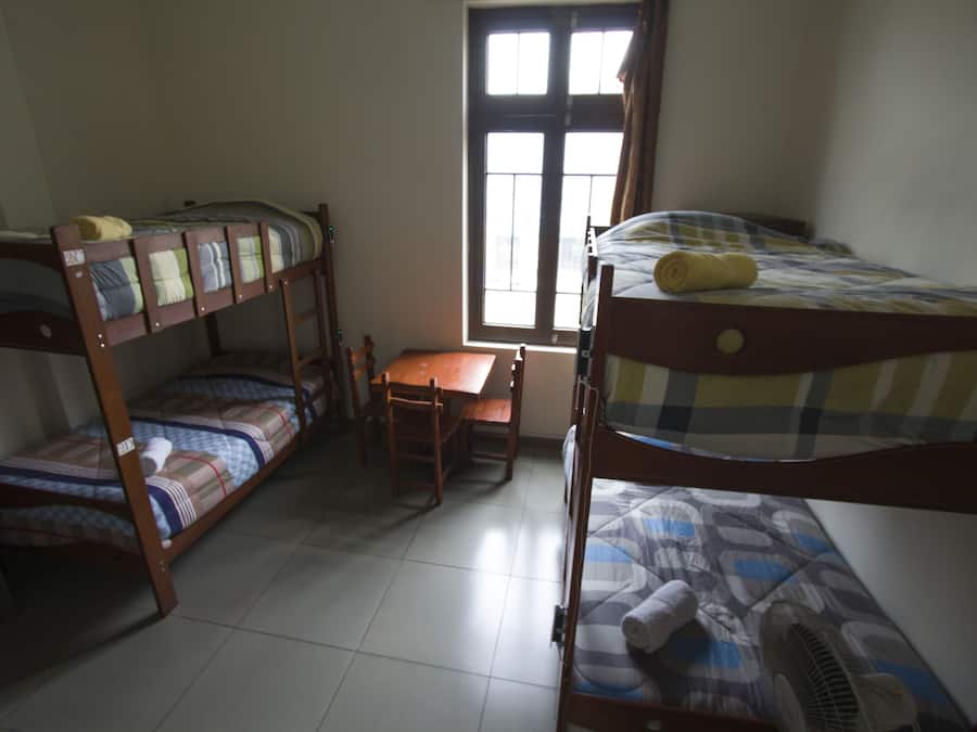 Quarto em dormitório com beliches, uma mesa pequena e uma janela.