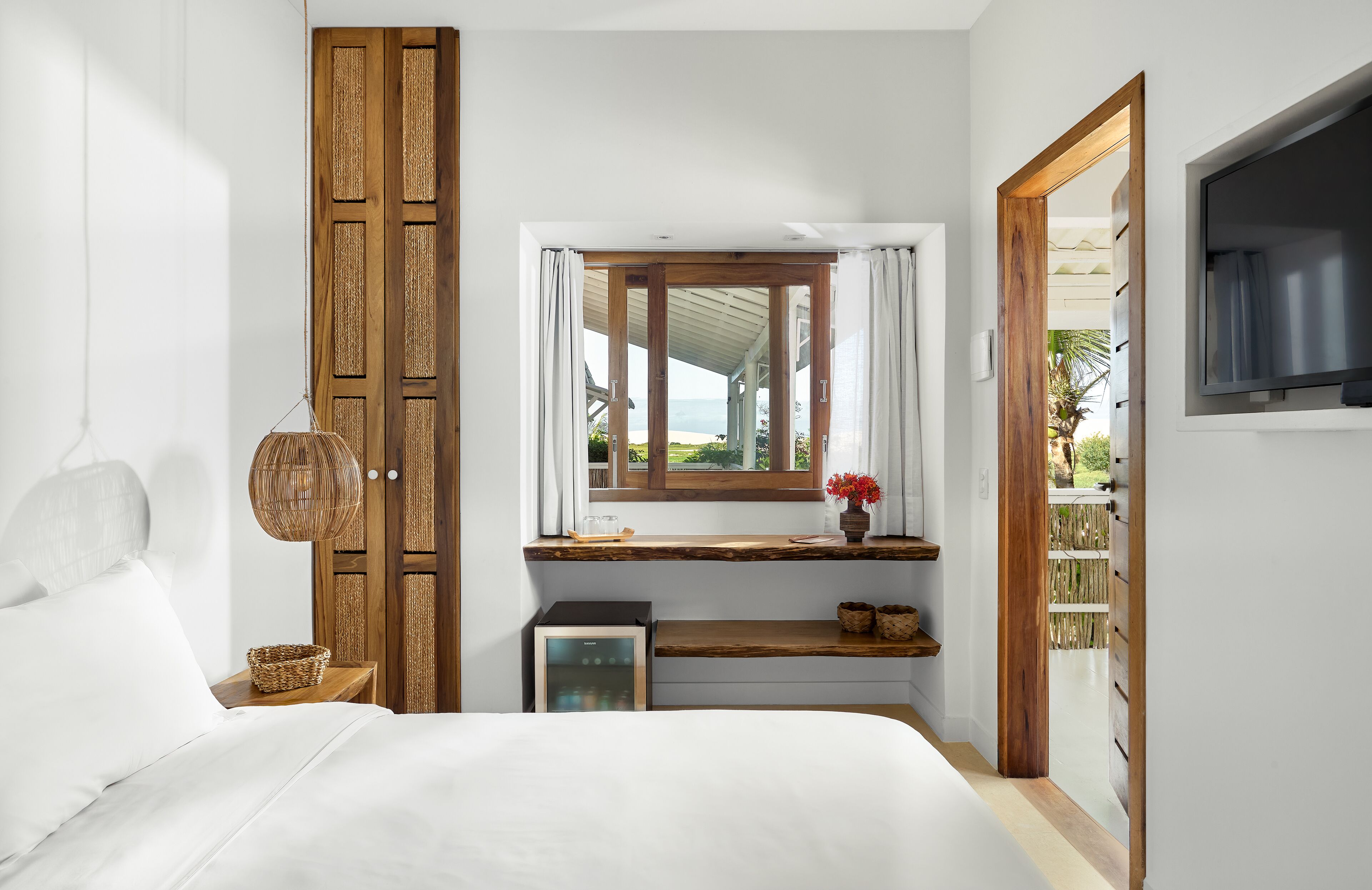 Quarto com cama, guarda-roupa de madeira, mesinha com cesta, janela com cortinas e televisão na parede.