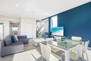 Uma sala de estar moderna com uma mesa de jantar de vidro, cadeiras brancas, um sofá cinza e uma grande TV de tela plana exibindo uma cena de praia.
