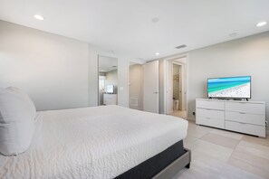 Quarto moderno com uma cama grande, televisão sobre uma cômoda branca e vista para uma praia pela janela.
