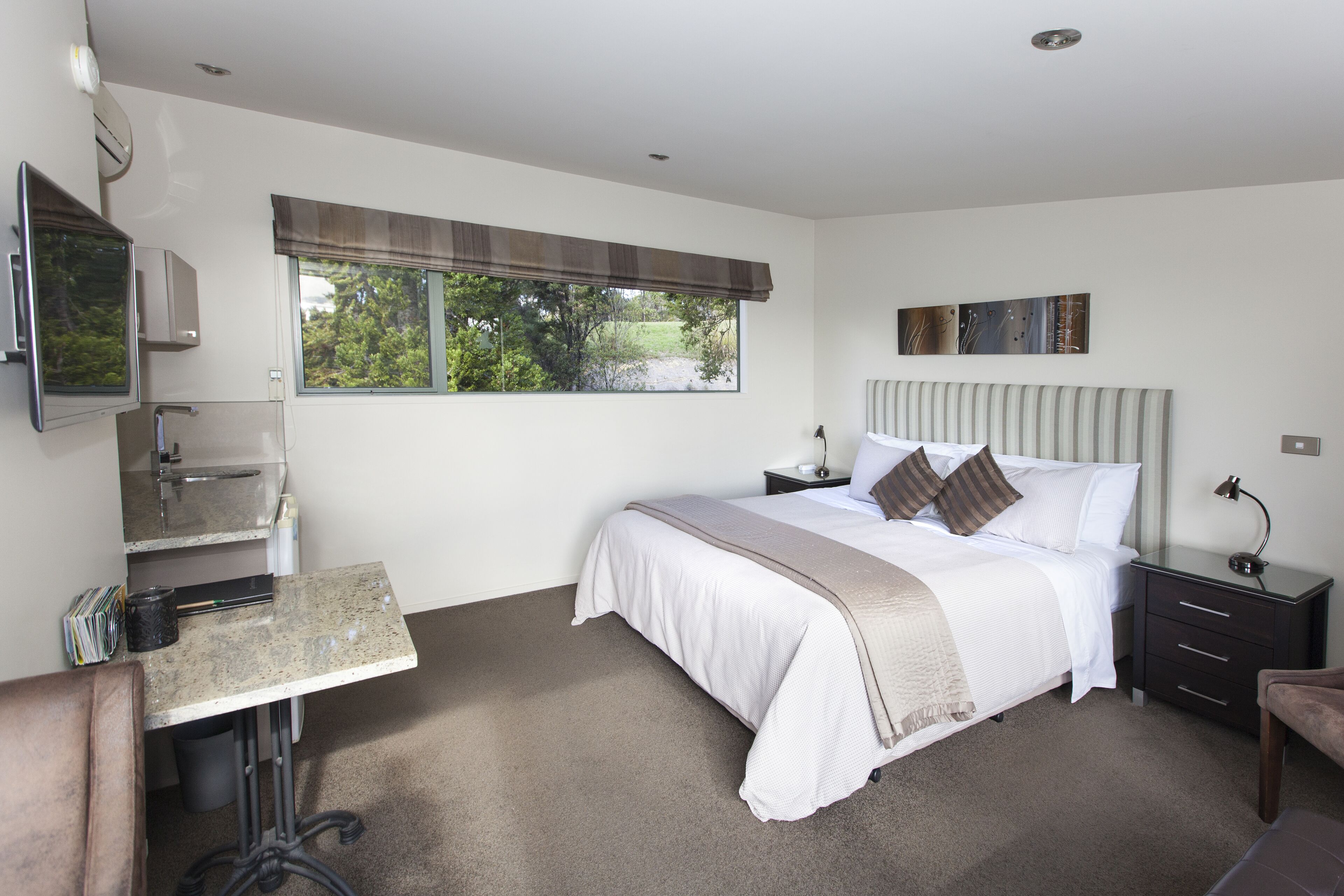 Quarto com cama, mesas de cabeceira, escrivaninha e janela com vista para o verde.