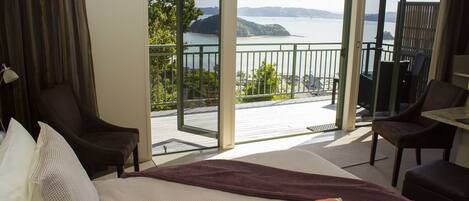 Quarto de hotel com uma cama grande, duas cadeiras, uma mesinha e uma varanda com vista para um lago e montanhas.