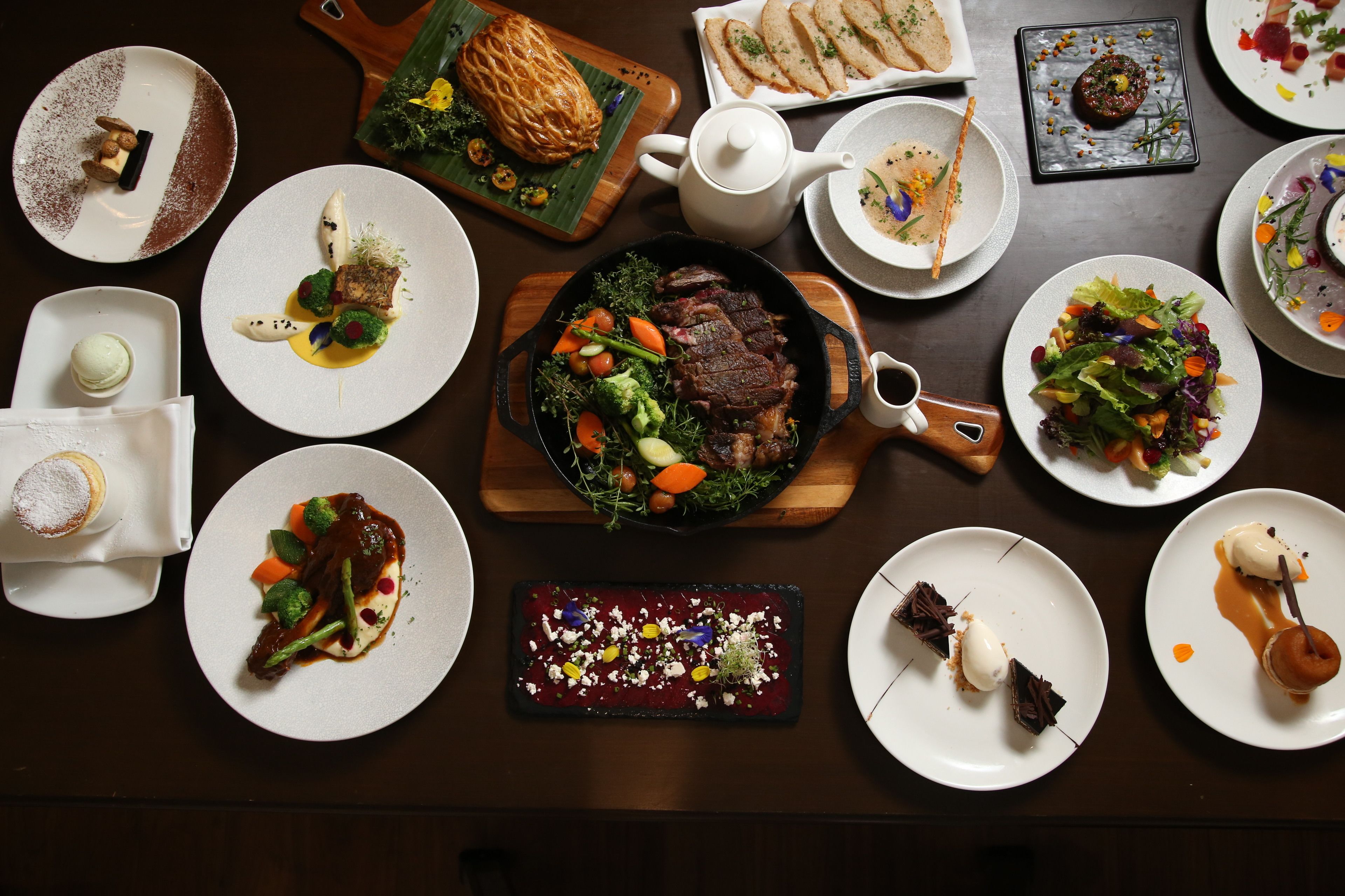 Diversos pratos sobre uma mesa de jantar, incluindo bife, legumes e sobremesas.