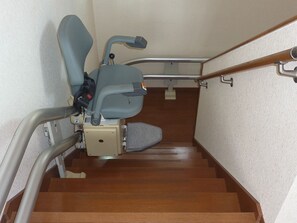 Escadaria com acesso para cadeiras de rodas, com corrimão, piso de madeira e painel de controle fixado na parede.