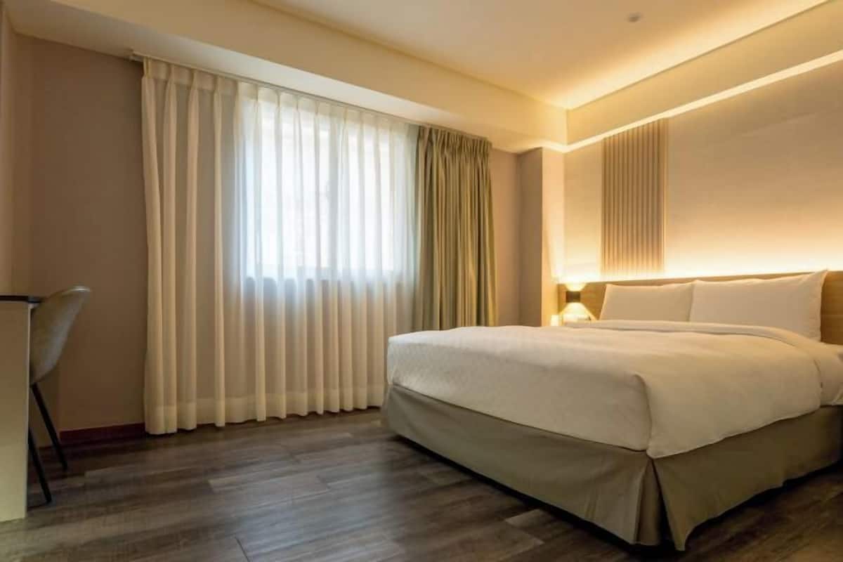 Quarto de hotel com cama, cortinas, escrivaninha e luminária fixada na parede.