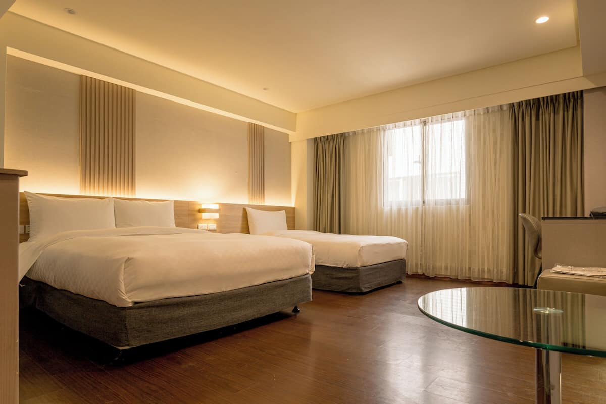 Quarto de hotel com duas camas, uma mesa de vidro e uma janela grande com cortinas.