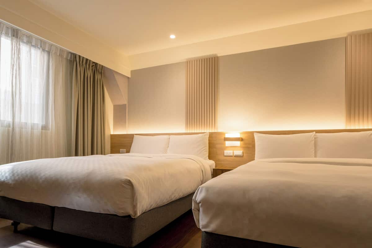 Quarto de hotel com duas camas, uma janela grande com cortinas e luminária fixada na parede.