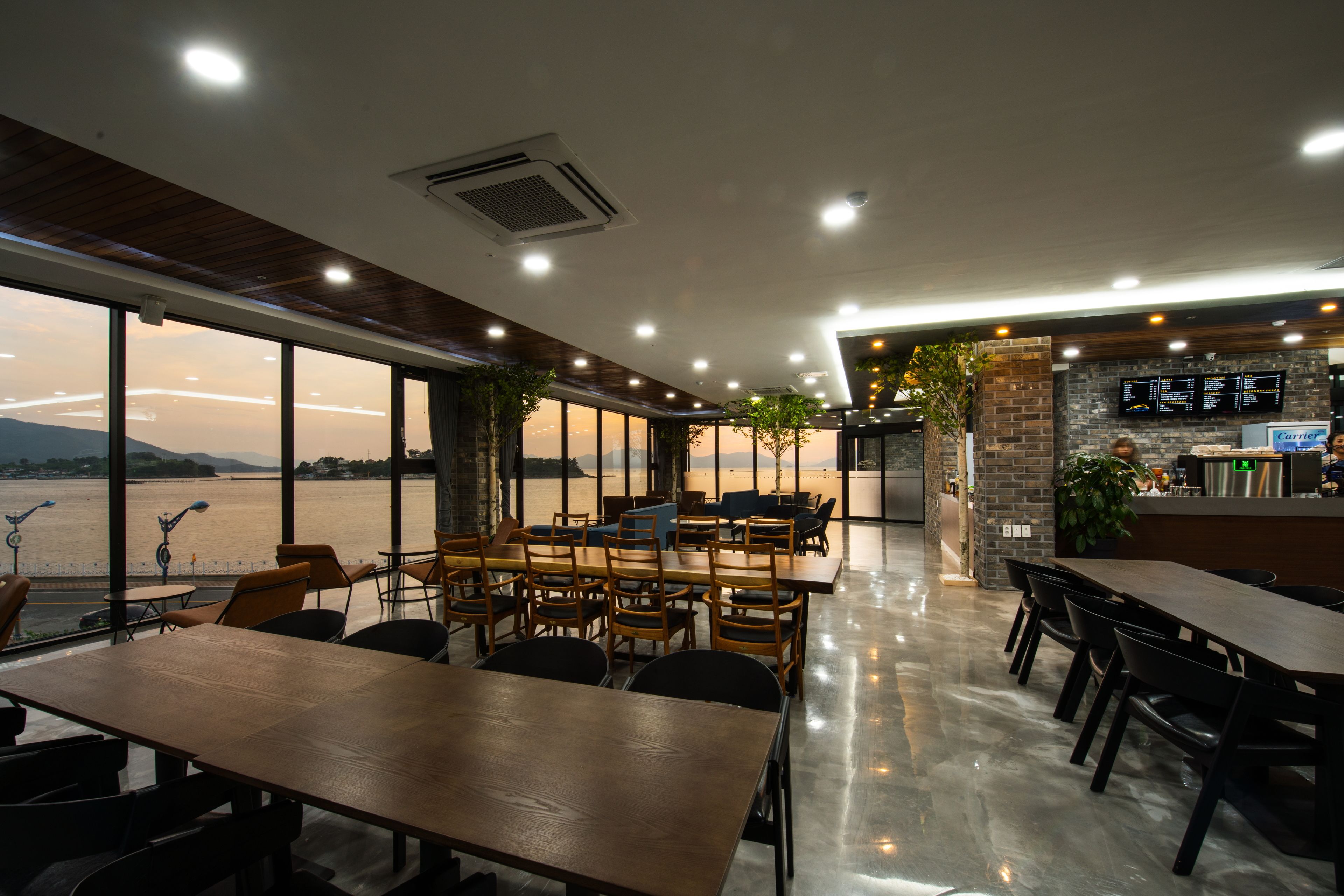 Restaurante com vista para um rio, mesas e cadeiras de madeira e uma parede de tijolos com um painel de cardápio digital.