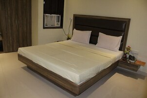 Uma cama grande com estrutura de madeira e roupa de cama branca, uma pequena mesa de cabeceira com um vaso de flores e um aparelho de ar condicionado fixado na parede.