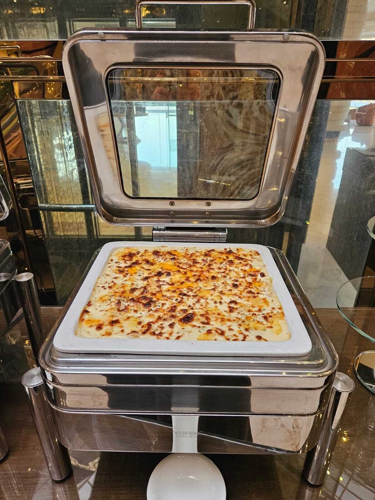 Disposição de buffet com um prato retangular para servir, contendo um prato assado, possivelmente lasanha, em recipiente metálico.