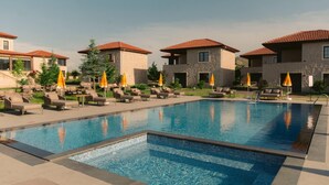 Piscina com espreguiçadeiras e guarda-sóis, cercada por edifícios de pedra com telhados de telha vermelha.