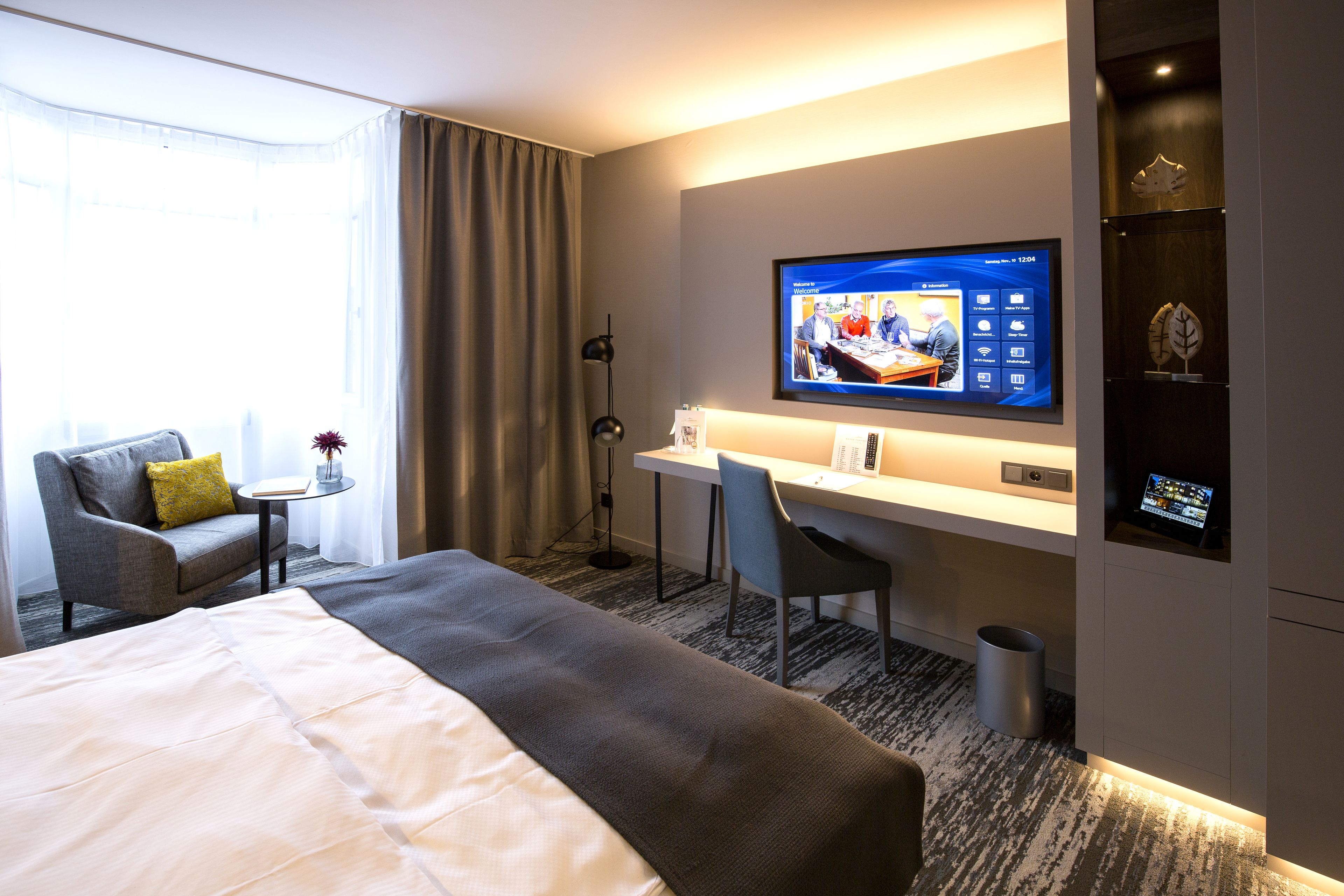 Quarto de hotel com cama, escrivaninha, cadeira, televisão e uma pequena mesa com um vaso.