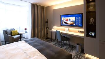 Quarto de hotel com cama, escrivaninha, cadeira, televisão e uma pequena mesa com um vaso.