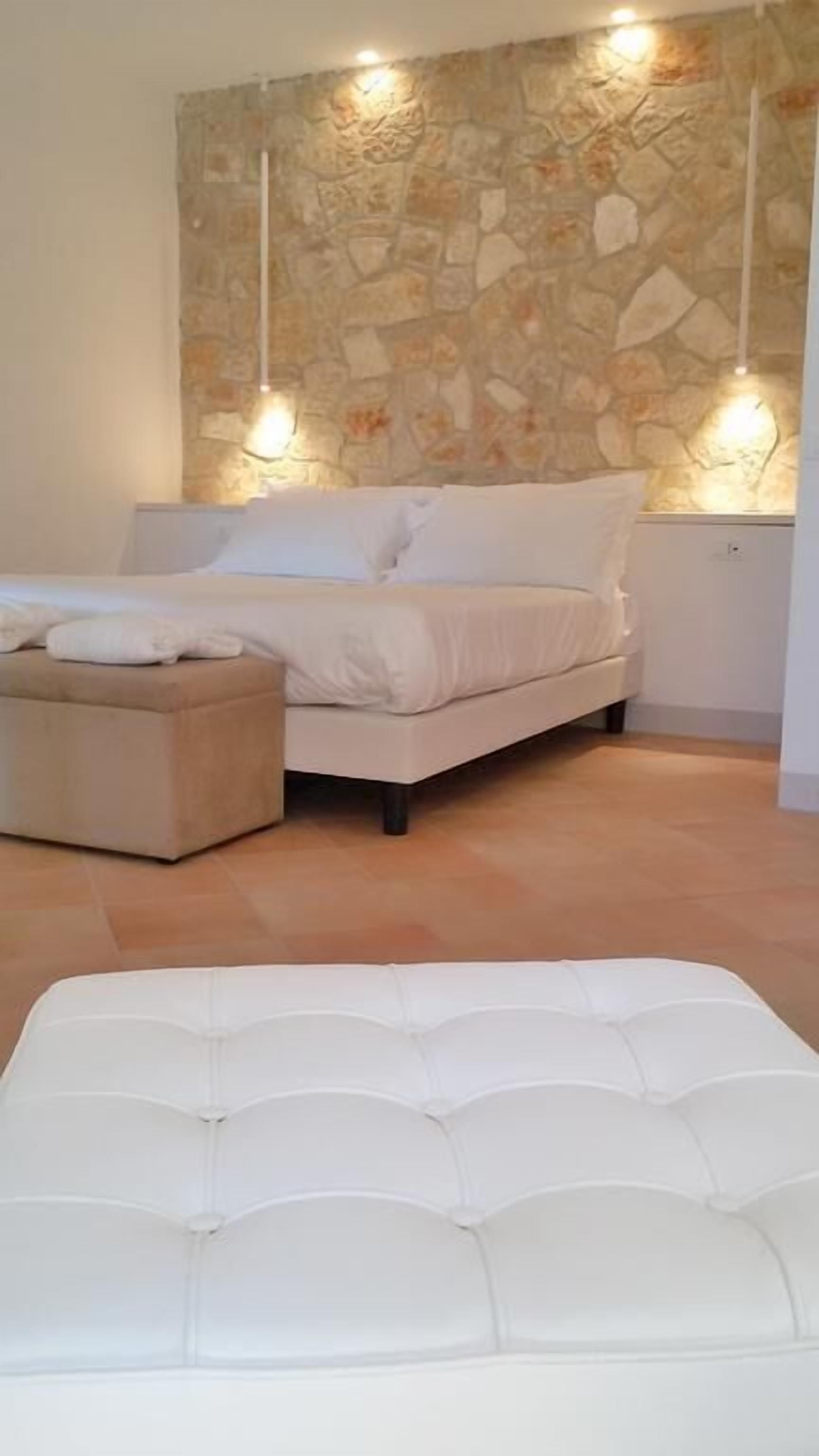 Quarto com parede de pedra, cama com roupa de cama branca, mesa de cabeceira e um pequeno banquinho.